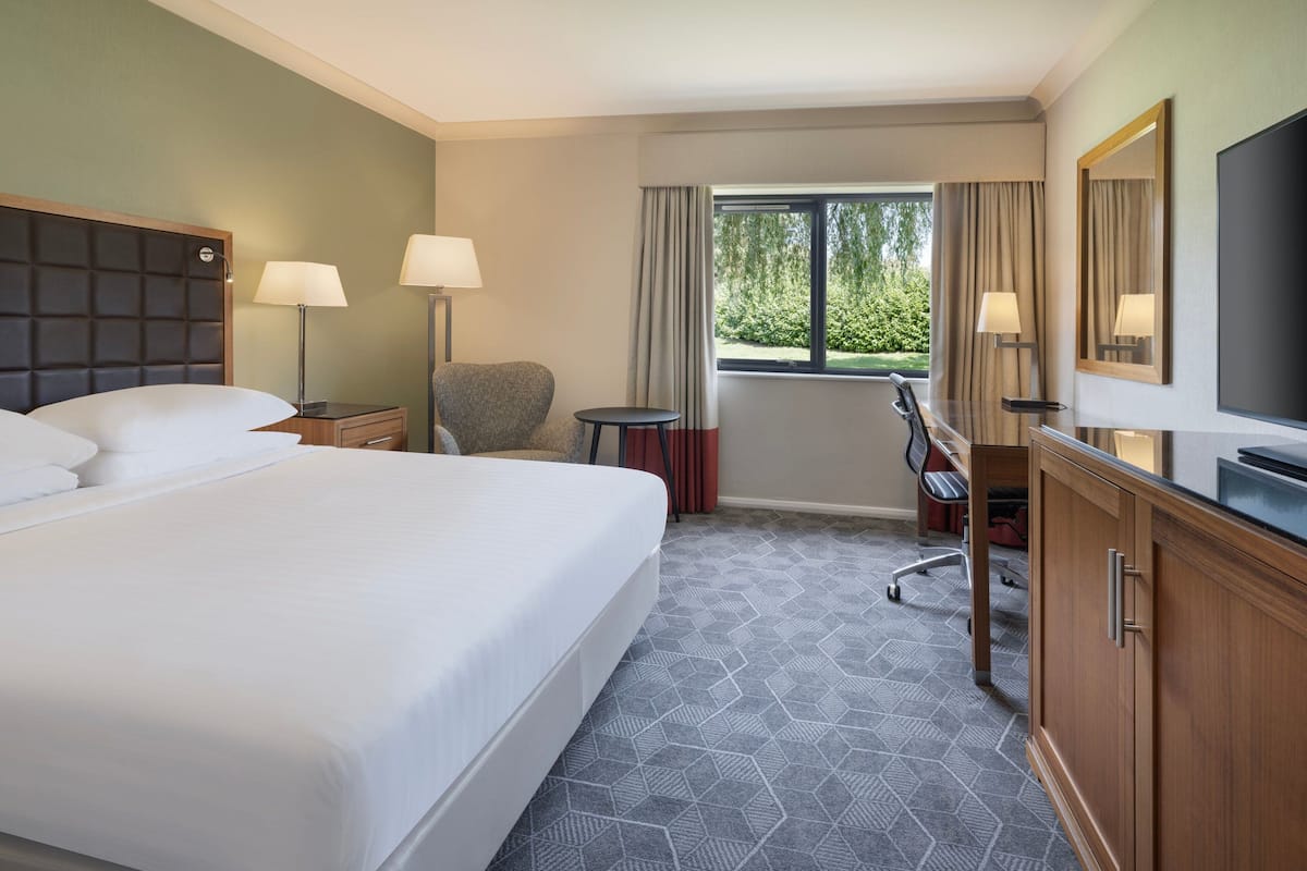 Quarto de hotel com uma cama grande, duas luminárias de cabeceira, uma escrivaninha com cadeira, televisão e janela com cortinas.