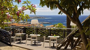 Terraço ao ar livre com mesas e cadeiras brancas, com vista para a costa, edifícios e o mar.