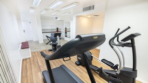Uma academia com esteiras e equipamentos elípticos, piso de madeira e paredes brancas.