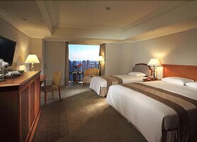 Quarto de hotel com duas camas, uma escrivaninha com televisão, uma cadeira e vista para a paisagem urbana.