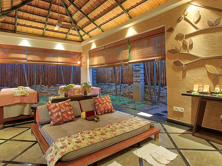 Um quarto amplo com teto alto, cama com colcha estampada, área de estar com almofadas, pequena mesa com planta e parede decorativa com motivos de folhas.