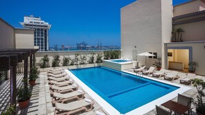 Piscina no terraço com espreguiçadeiras e plantas em vasos, com vista para um porto e suas guindastes.