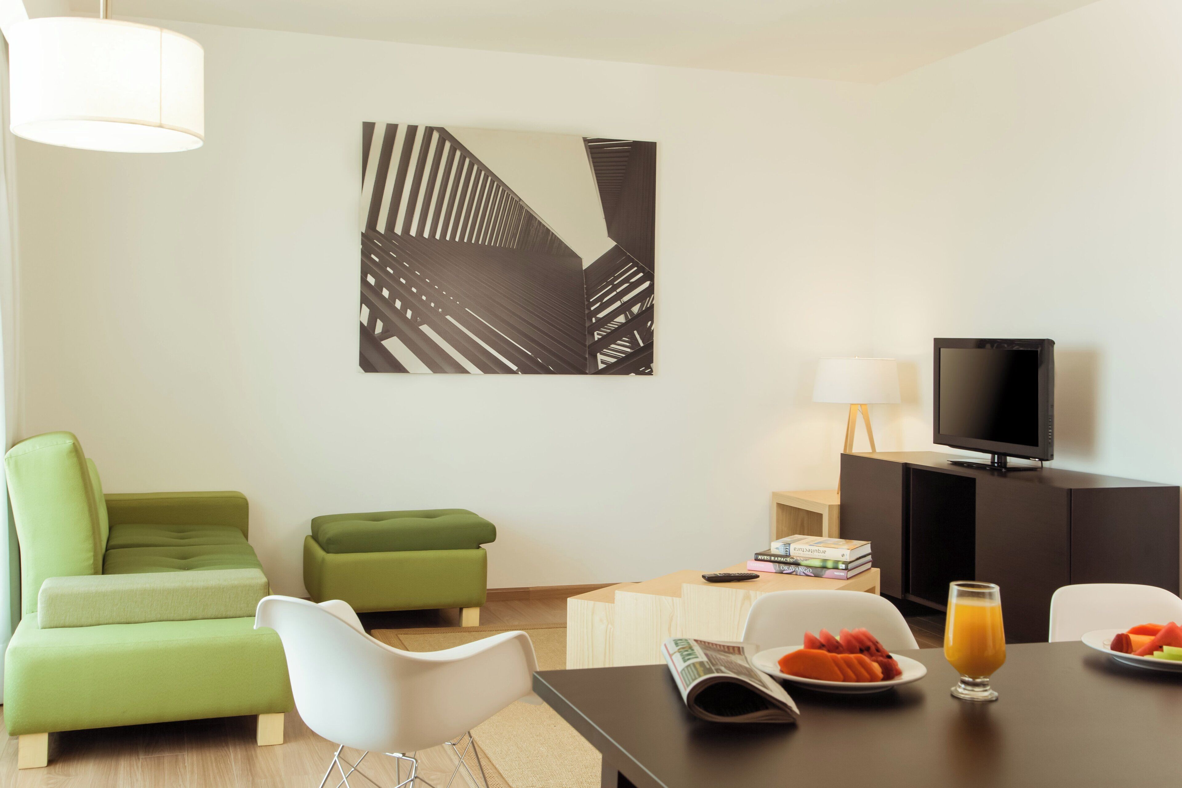 Uma sala de estar moderna com um sofá verde, uma cadeira branca, uma mesa de jantar com uma revista, uma travessa de frutas e uma TV sobre um móvel.