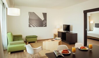 Quarto de hotel com cama, televisão, escrivaninha com luminária, sofá verde, cadeira branca e mesa de jantar com fruta e suco.