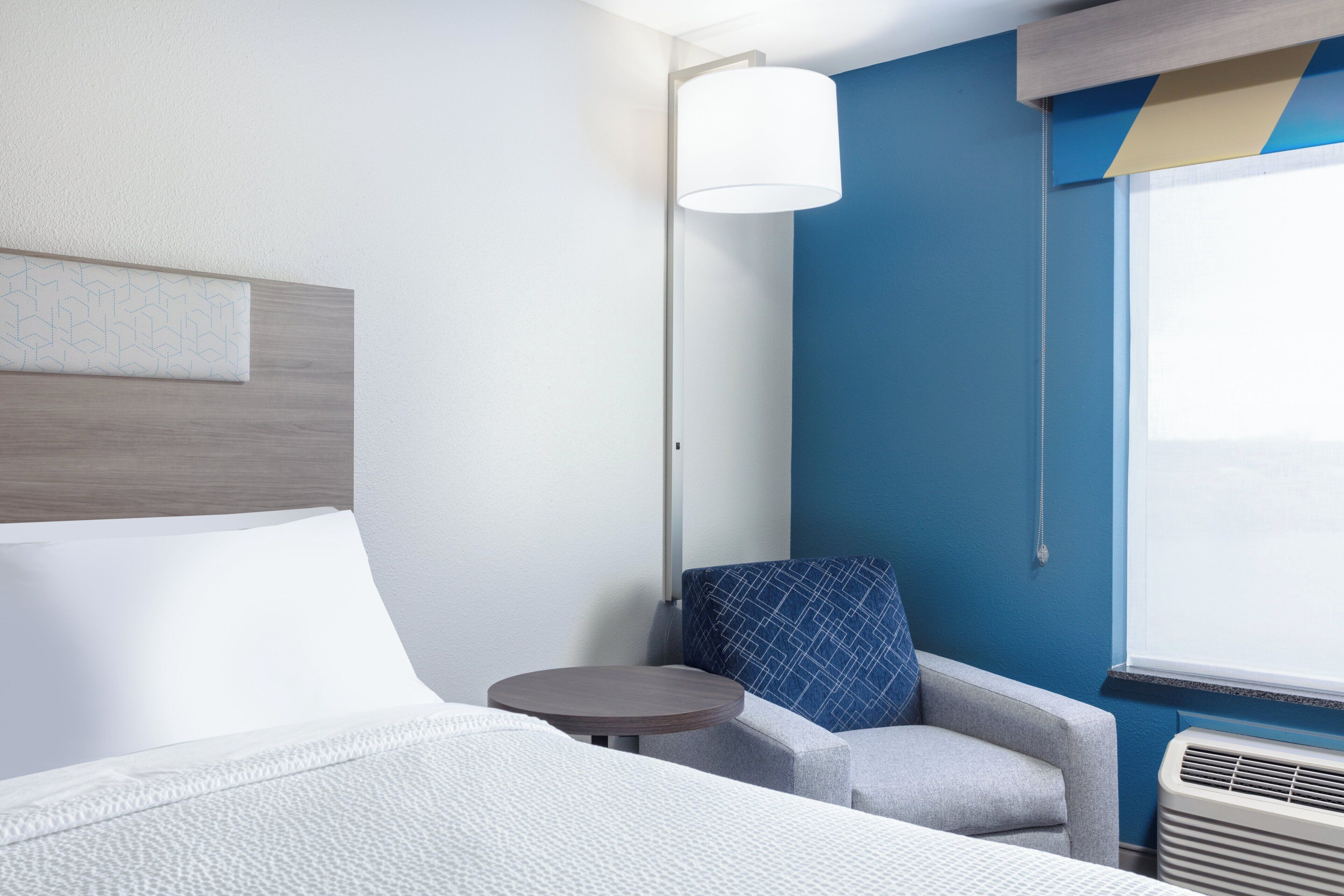 Quarto de hotel com cama, mesa redonda pequena, cadeira, abajur e janela.