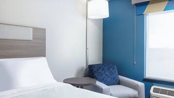 Quarto de hotel com cama, mesa redonda pequena, cadeira, abajur e janela.