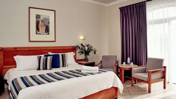 Quarto de hotel com cama grande, cabeceira de madeira, quadro emoldurado na parede, pequena mesa com vaso de flores, cadeira e janela com cortinas roxas.