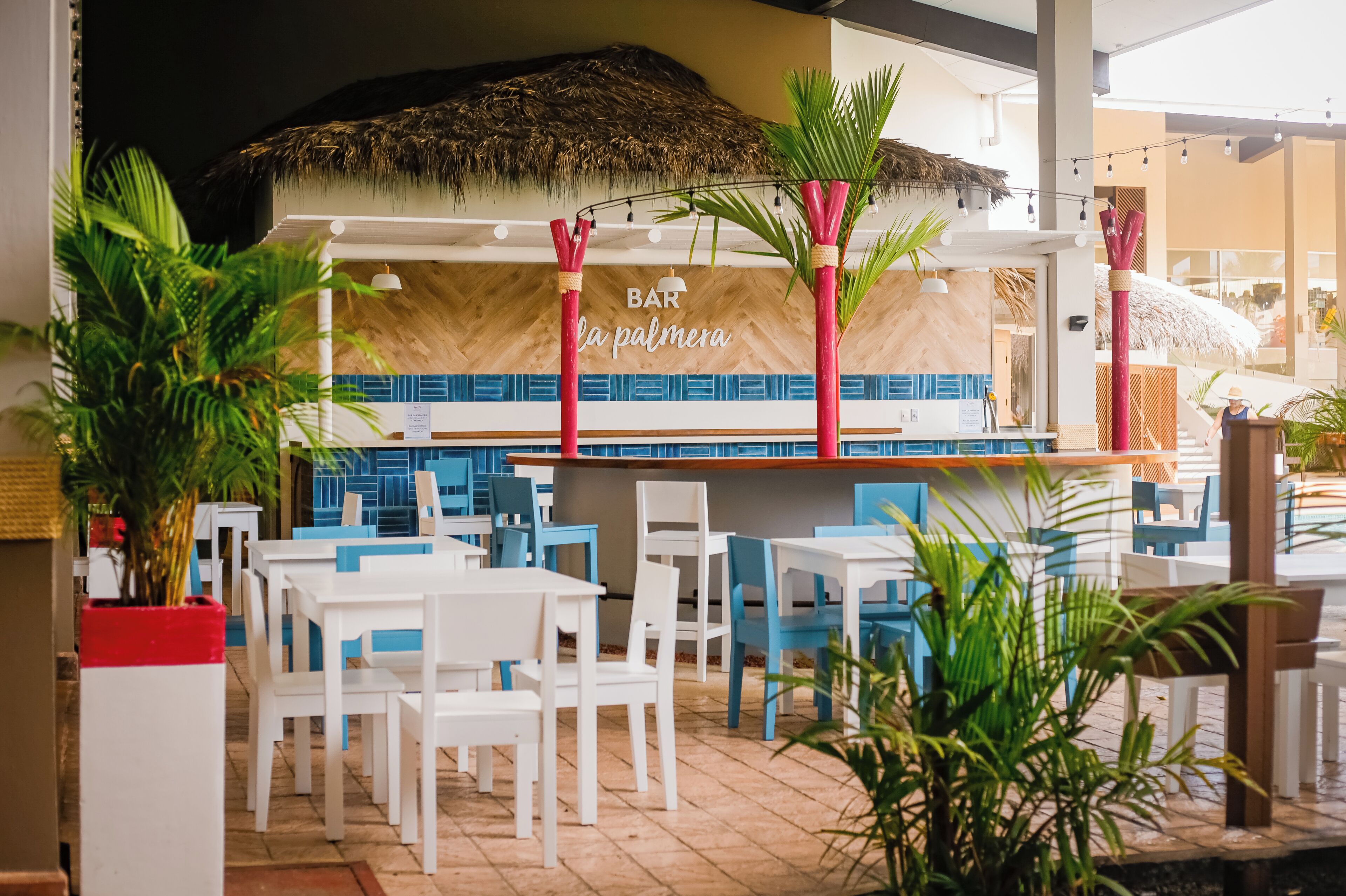 Uma área de refeições ao ar livre com mesas e cadeiras brancas e azuis, um bar com cobertura de palha e plantas tropicais.