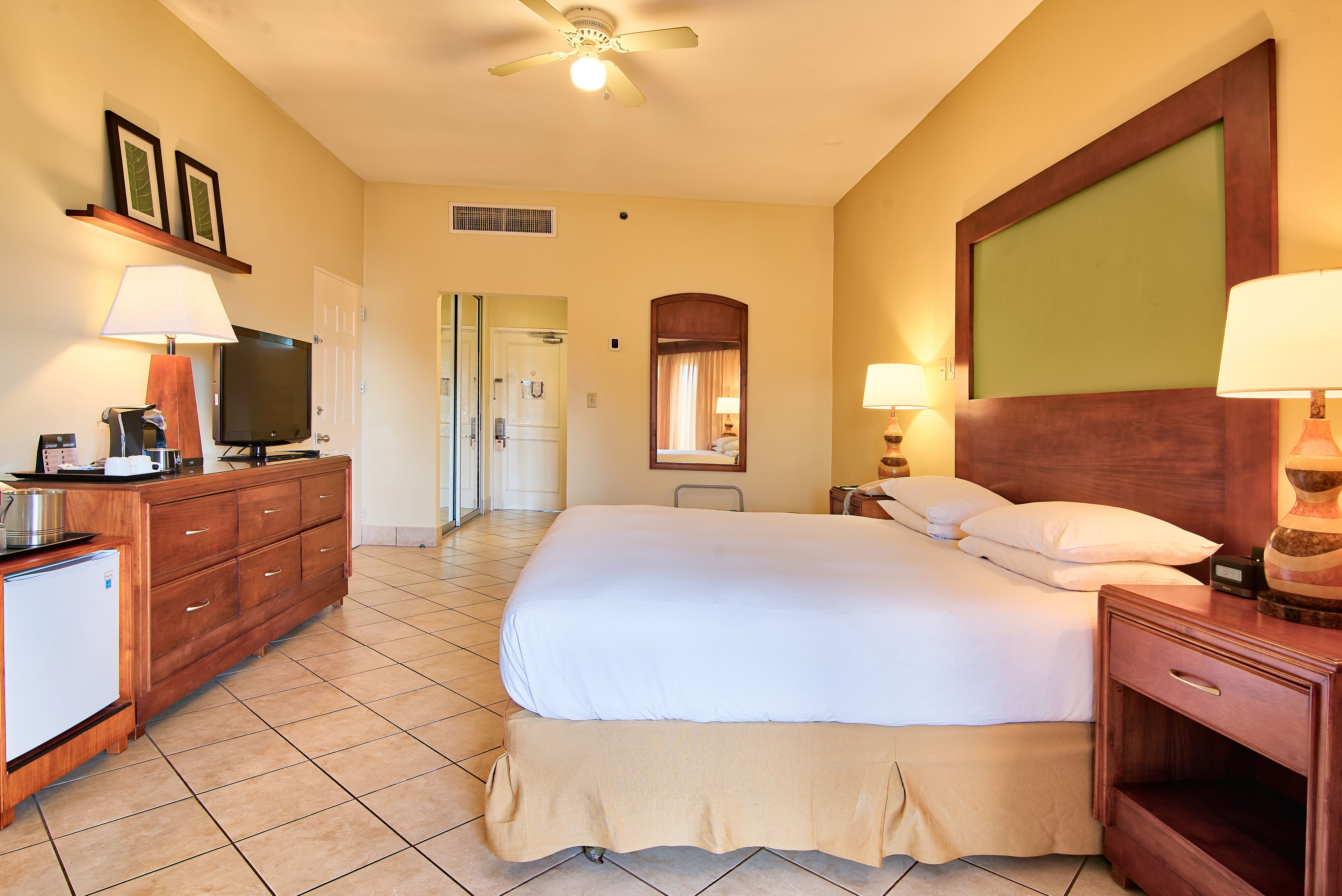 Quarto de hotel com cama, escrivaninha, televisão, mini-geladeira e ventilador de teto.