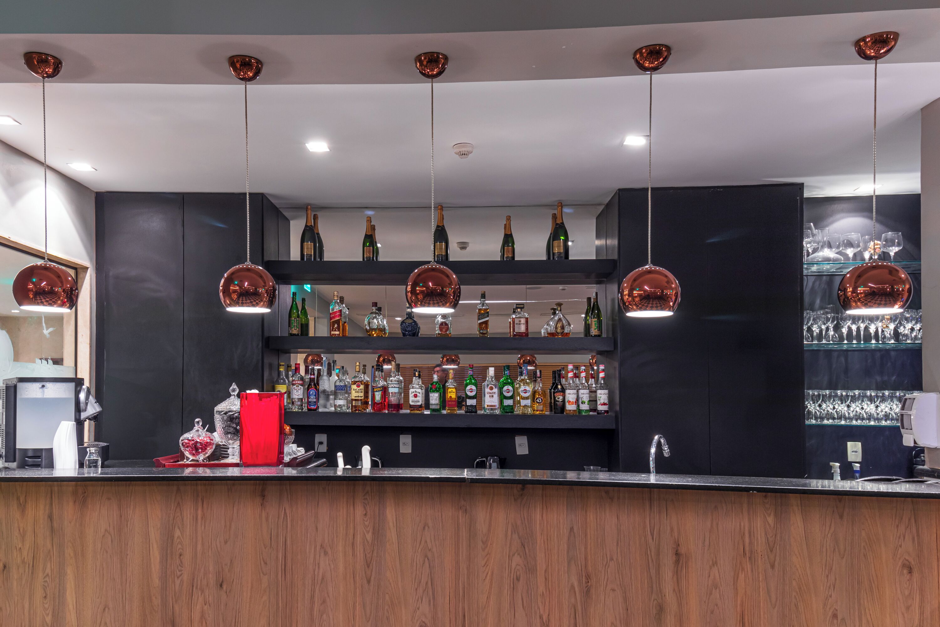 Uma área de bar com balcão de madeira, luminárias pendentes em cobre, prateleiras com garrafas de bebidas e uma vitrine para copos de vinho.