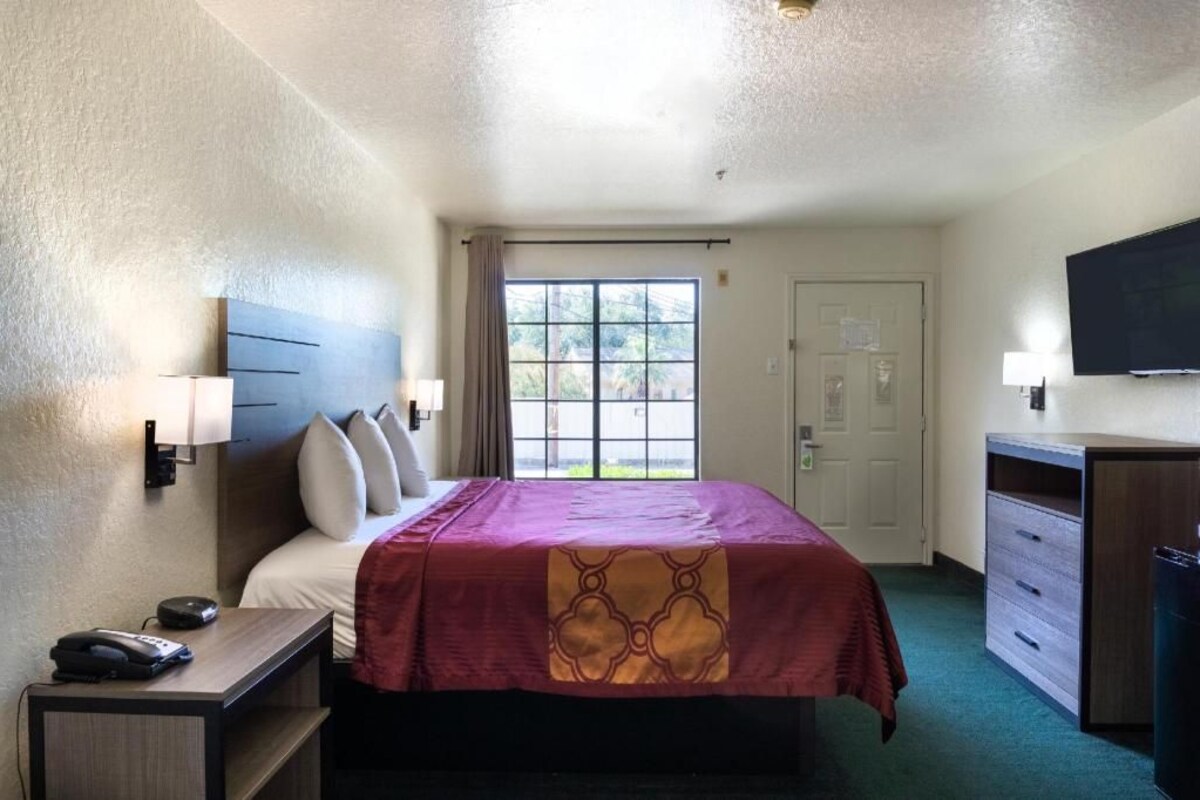 Quarto de hotel com cama, abajures, escrivaninha com telefone, televisão e janela com vista.