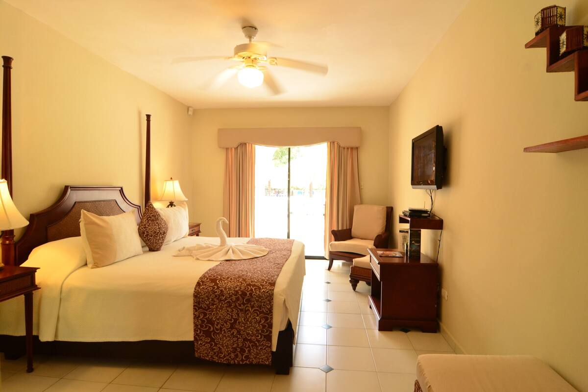 Quarto de hotel com uma cama grande, uma escrivaninha, uma cadeira, uma televisão e um ventilador de teto.