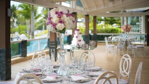 Mesa de banquete montada para um evento formal, com um arranjo floral, toalhas brancas e copos de vidro transparente.