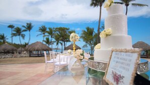 Bolo de casamento sobre uma mesa de vidro, com paisagem tropical ao fundo.