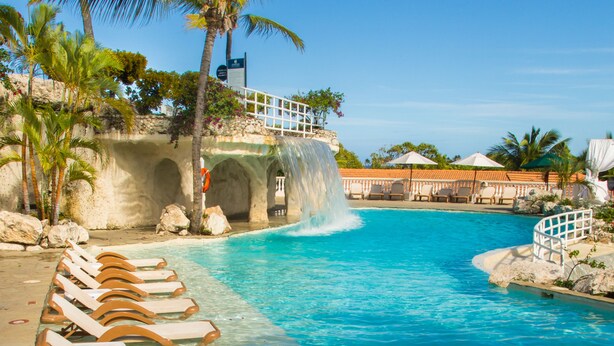 Piscina com águas cristalinas, espreguiçadeiras, palmeiras e uma cascata.