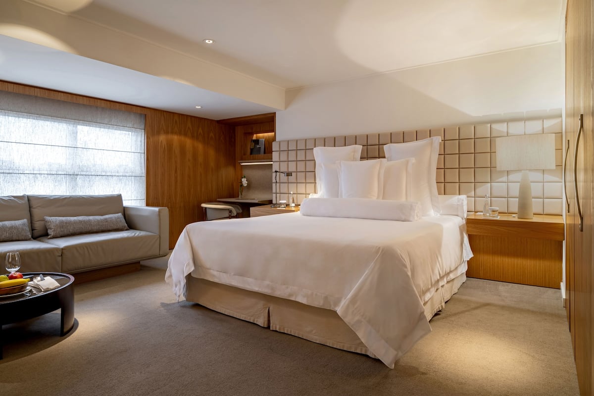 Quarto de hotel moderno com uma cama grande, um sofá, uma mesa de centro com frutas e vista para a cidade.