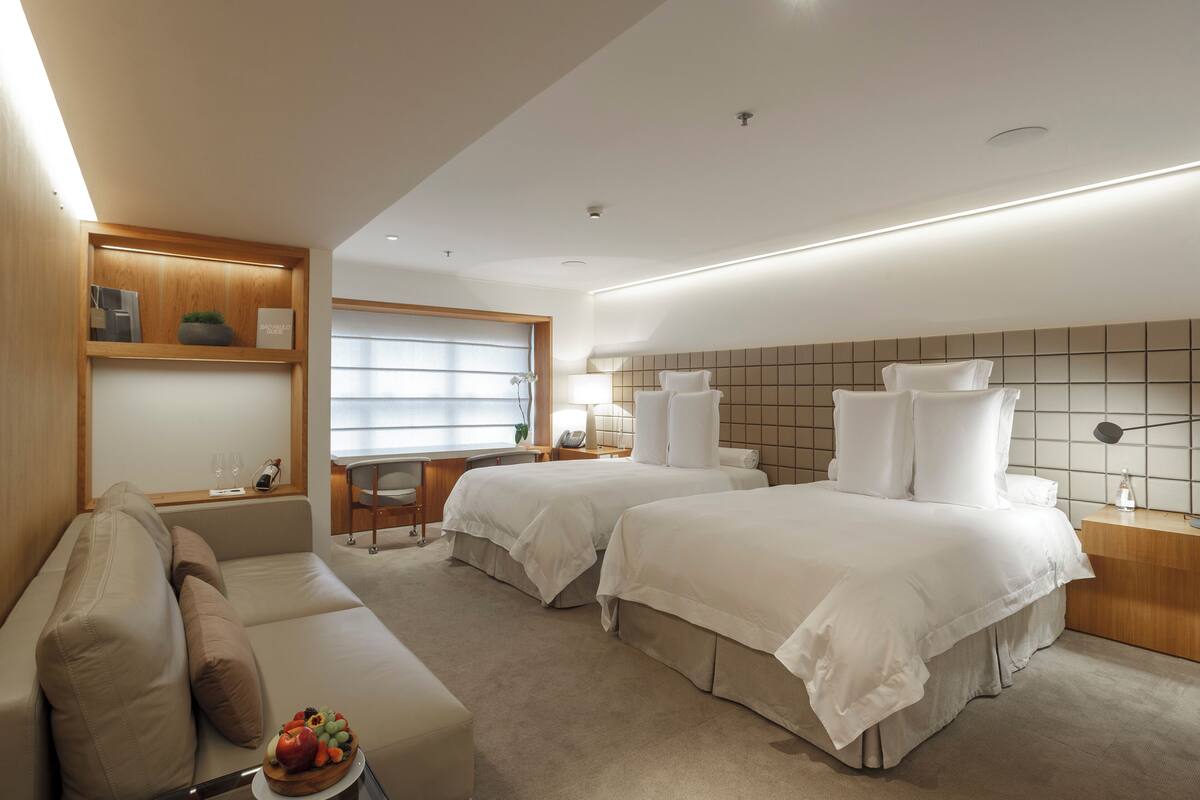 Quarto de hotel moderno com duas camas, um sofá, uma pequena mesa com frutas, uma escrivaninha e uma janela com persianas.