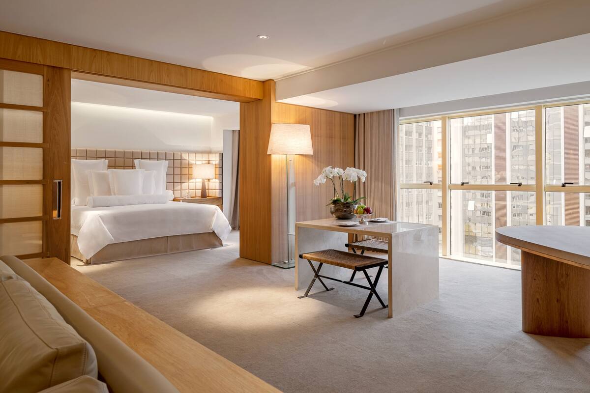 Quarto de hotel moderno com uma cama grande, uma escrivaninha com uma luminária, uma mesinha com flores e vista da cidade através de amplas janelas.
