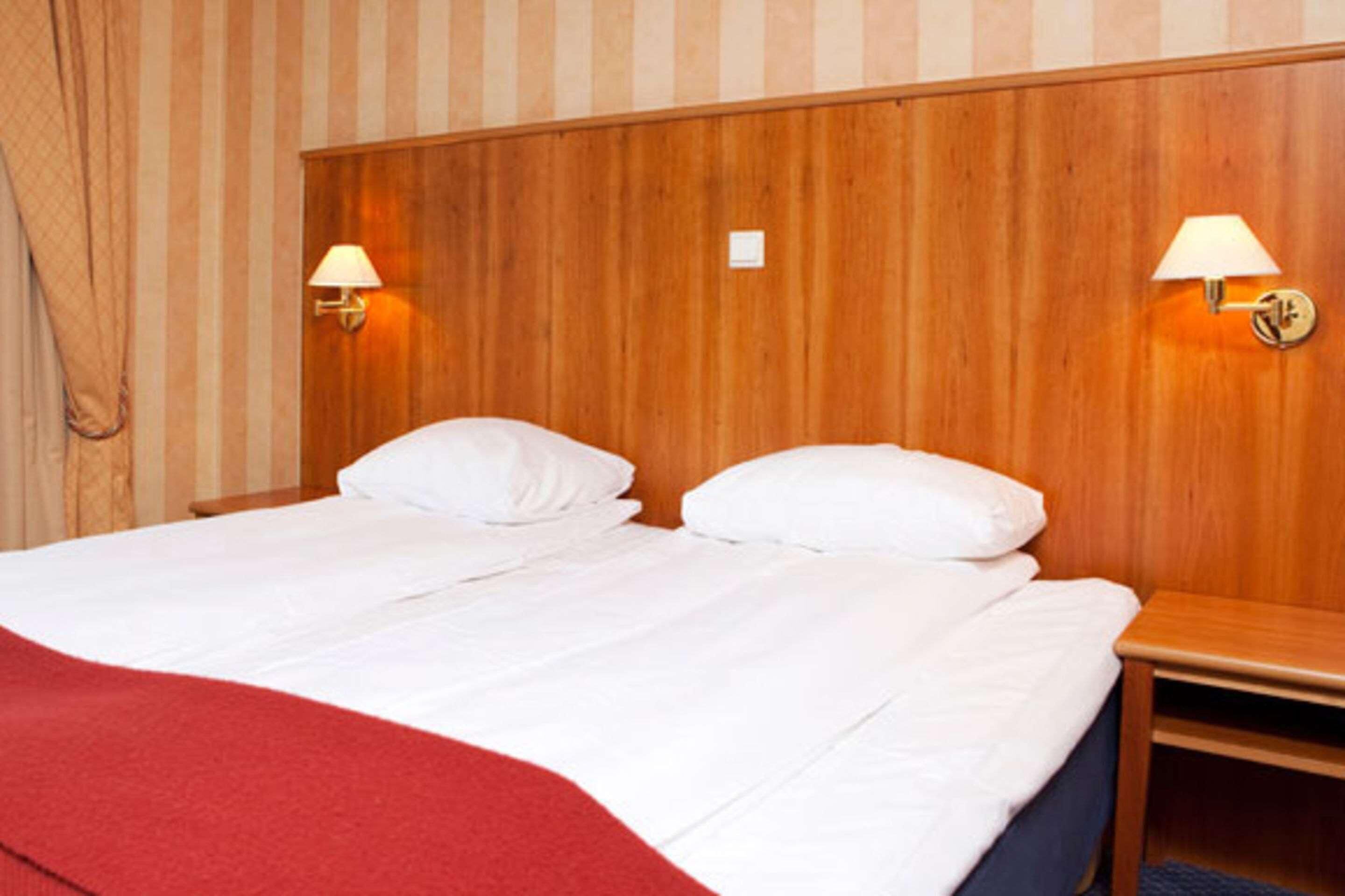 Quarto de hotel com cabeceira de madeira, duas luminárias de cabeceira, cama com lençóis brancos e um cobertor vermelho.