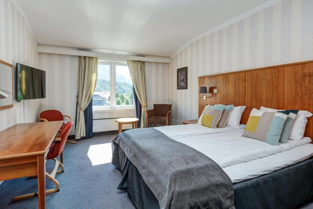 Quarto de hotel com uma cama, escrivaninha, cadeira, televisão e janela com cortinas, e vista para as montanhas.