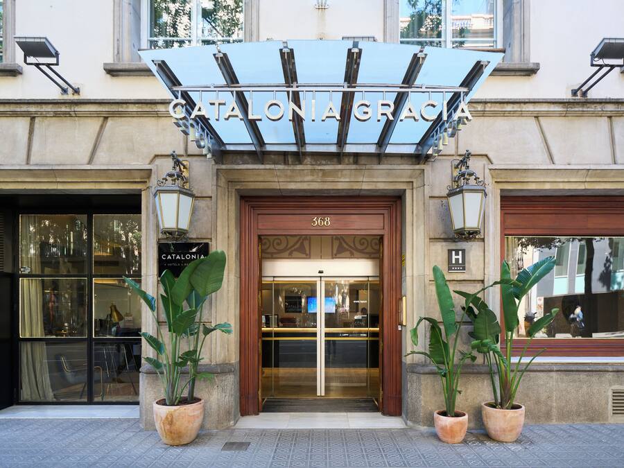 Entrada de um hotel com porta de vidro, plantas em vasos e uma placa acima da porta.
