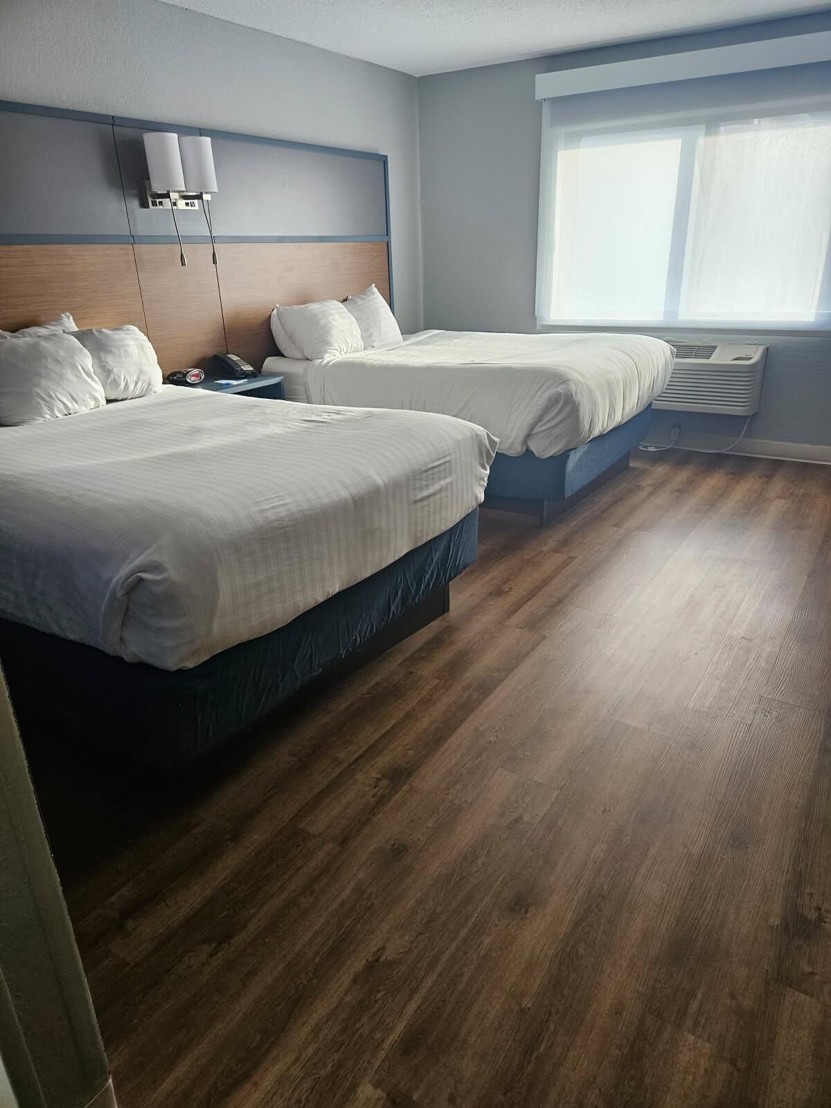 Quarto de hotel com duas camas, chão de madeira, cabeceira com luminária e janela com persianas.
