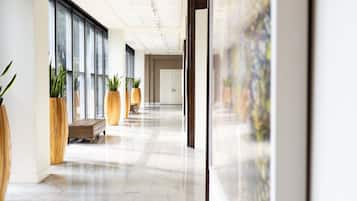 Corredor moderno de hotel com grandes janelas, plantas em vasos e piso de mármore.