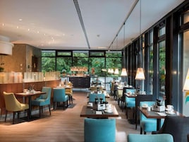 Um restaurante moderno com área de bar, mesas e cadeiras, e amplas janelas com vista para áreas verdes.