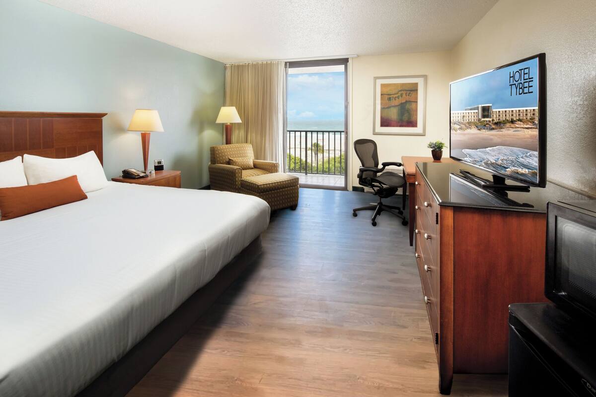 Quarto de hotel com cama, duas cadeiras, uma mesa de trabalho, televisão e varanda com vista.