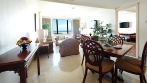 Quarto de hotel com área de jantar, sofá, televisão e vista para o mar.