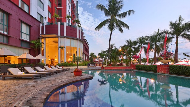Área de piscina de hotel com espreguiçadeiras e guarda-sóis, cercada por palmeiras e um edifício com fachada vermelha.
