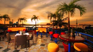 Terraço com mesas e cadeiras, com vista para um corpo d'água e um pôr do sol ao fundo.