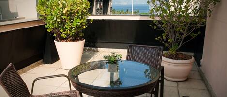 Uma mesa de vidro redonda com cadeiras de vime numa varanda com plantas em vasos e vista para o mar.