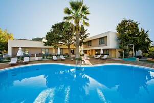 Piscina com espreguiçadeiras e guarda-sóis, cercada por edifícios modernos e árvores.