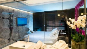 Quarto de hotel moderno com uma cama grande, banheiro com pia e espelho, e uma TV fixada na parede.