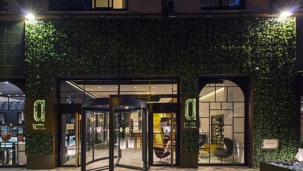 Entrada de hotel com porta giratória, portas de vidro e parede coberta por folhagem verde.