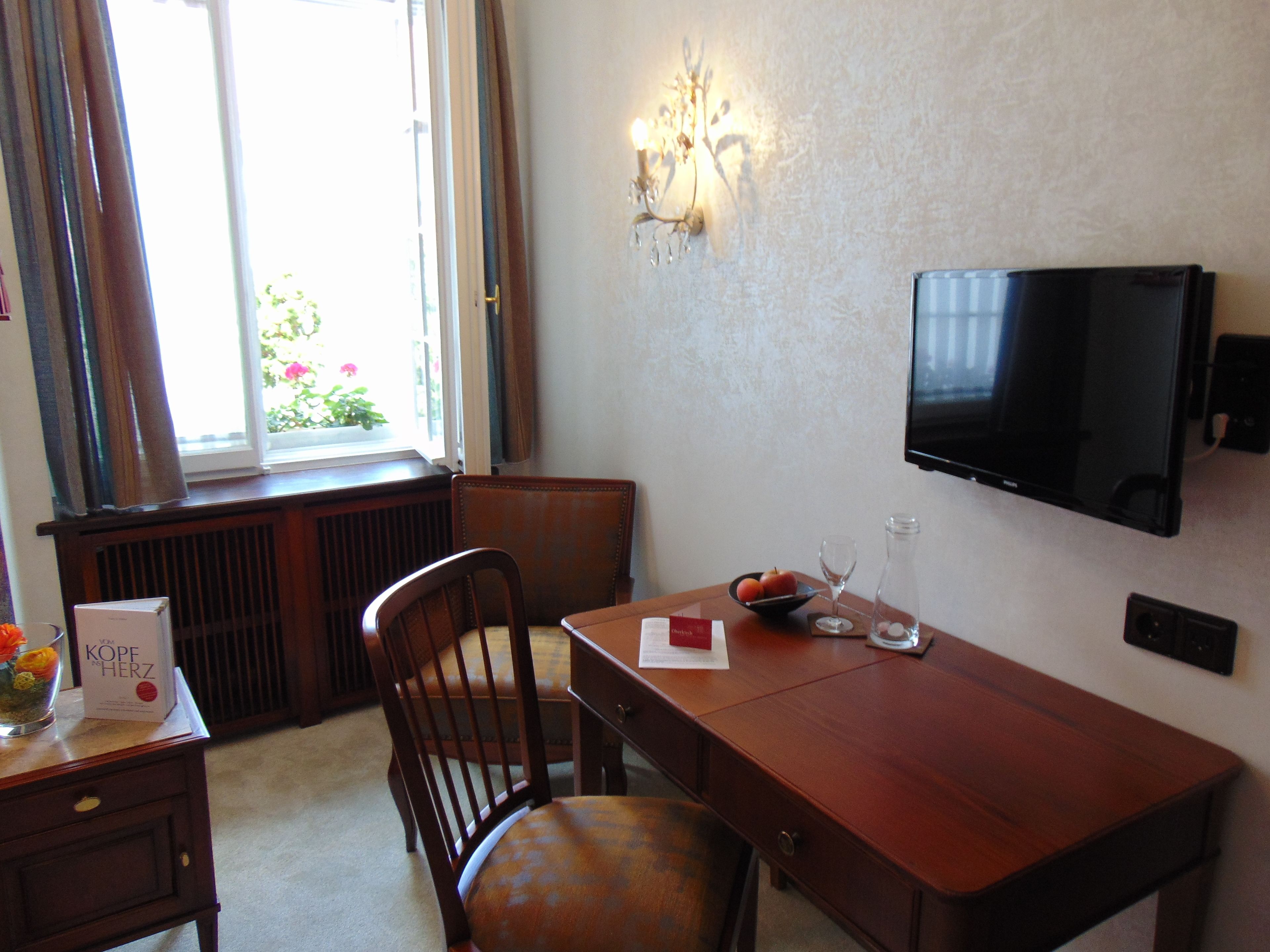 Quarto de hotel com escrivaninha de madeira, uma cadeira, televisão presa à parede e janela com cortinas.