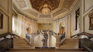 Uma escadaria imponente com grades elaboradas e um candelabro, que leva a um espaço com pé-direito alto e detalhes decorativos no teto.