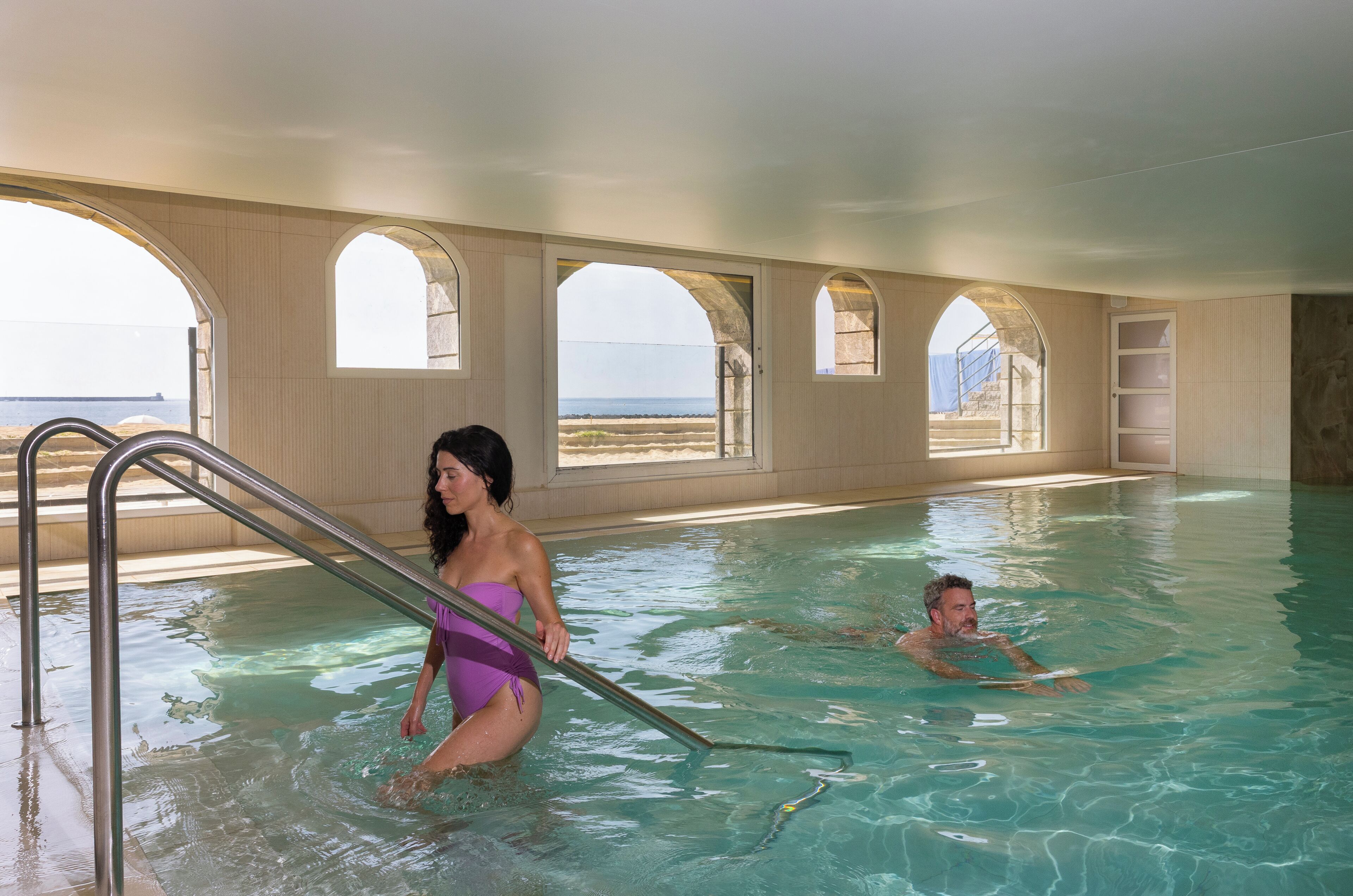 Piscina interior com janelas em arco, escada visível e vista clara para o mar através das janelas.