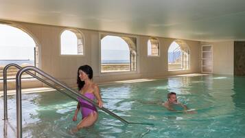 Piscina interior com janelas em arco, escada visível e vista clara para o mar através das janelas.