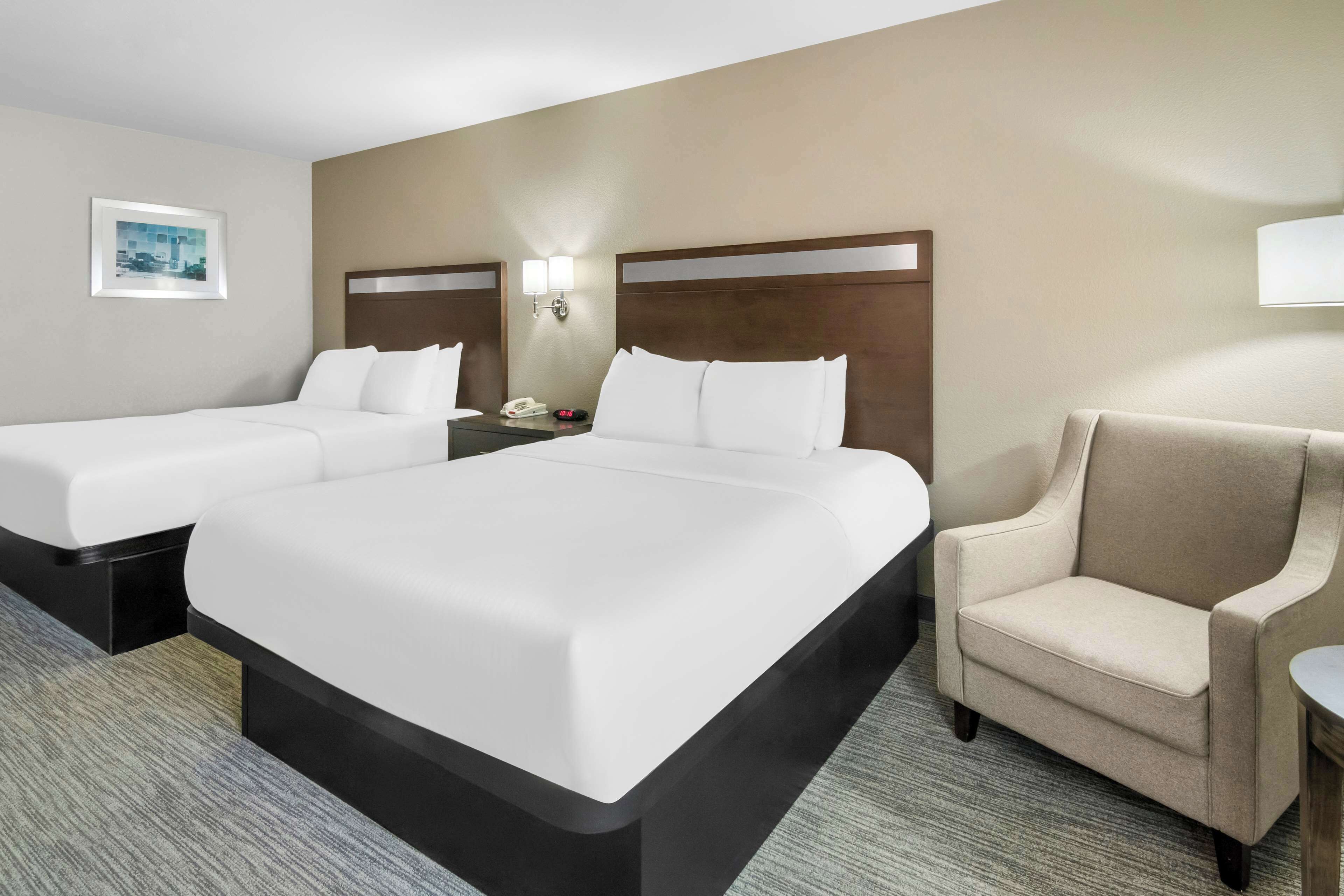 Quarto de hotel com duas camas de solteiro, uma cama de casal, uma cadeira, mesa de cabeceira com abajur e um quadro na parede.