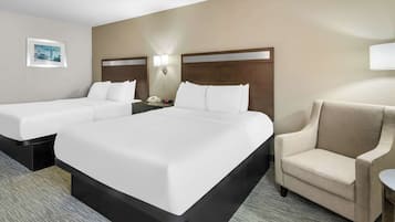 Quarto de hotel com duas camas de solteiro, uma cama de casal, uma cadeira, mesa de cabeceira com abajur e um quadro na parede.