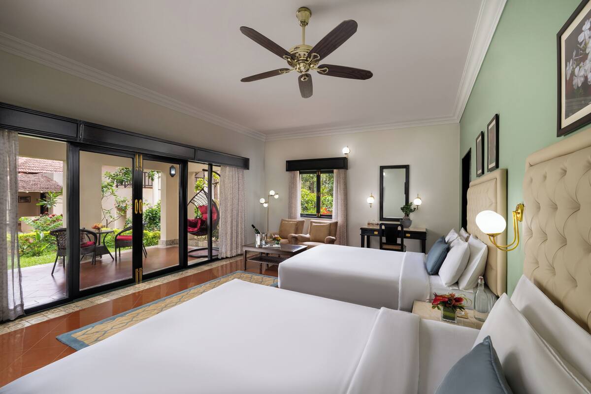 Quarto de hotel com duas camas, um ventilador de teto, uma janela grande e vista para o jardim.