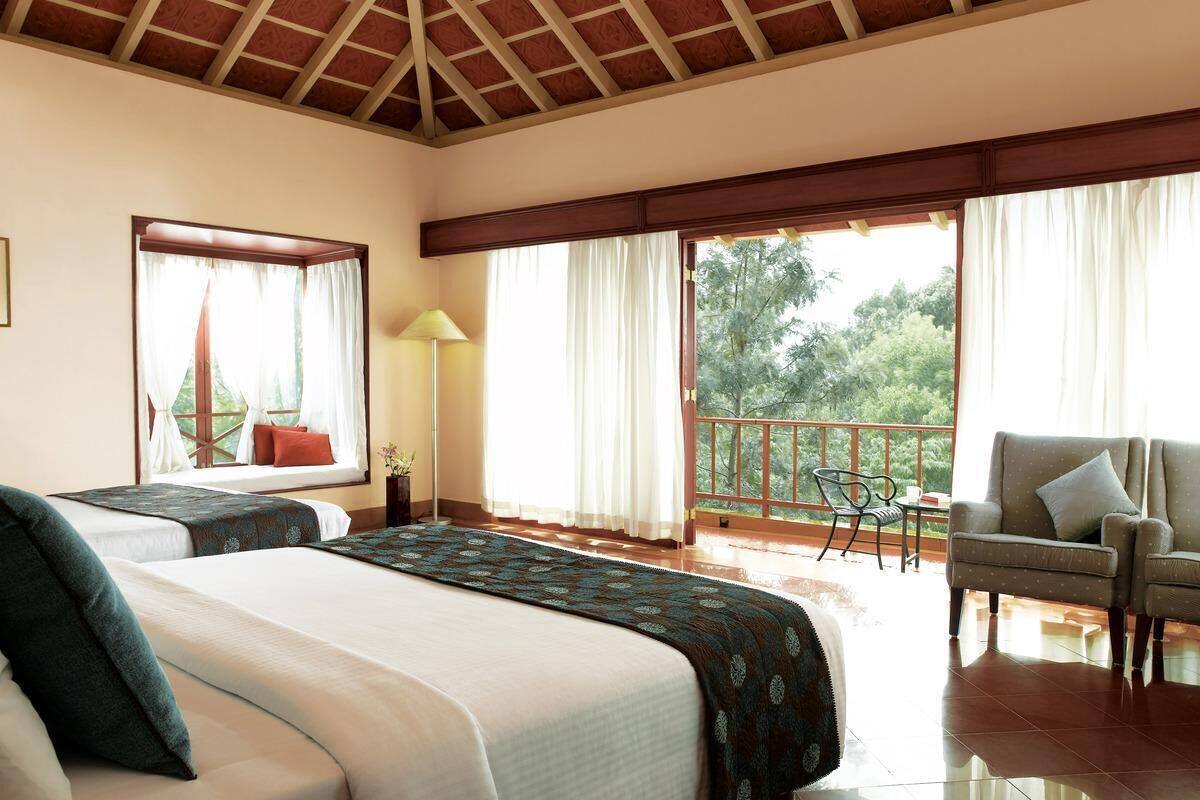 Quarto de hotel com duas camas, varanda com uma cadeira e vista para o verde.