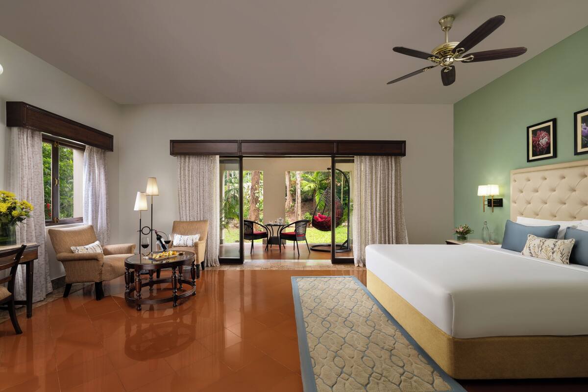 Quarto de hotel com uma cama grande, um ventilador de teto, área de jantar com mesa e cadeiras, e vista para o exterior.