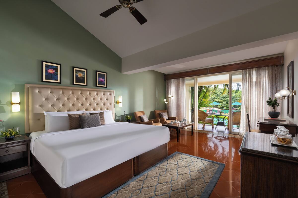 Quarto de hotel com uma cama grande, ventoinha no teto, vista para o exterior e área de refeições com mesa e cadeiras.