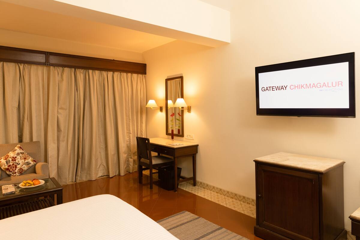 Quarto de hotel com cama, escrivaninha, cadeira, televisão e guarda-roupa.