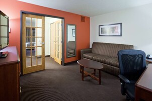 Quarto de hotel com secretária de madeira, uma mesinha redonda, sofá estampado, cadeira de escritório azul e porta corrediça para uma casa de banho.
