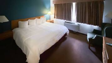 Quarto de hotel com uma cama grande, uma escrivaninha com cadeira, uma luminária e uma janela com cortinas.
