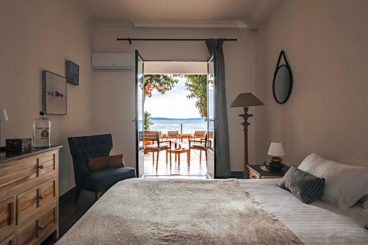 Quarto com cama, uma cadeira, cômoda, abajur e vista para o mar através de uma porta aberta.